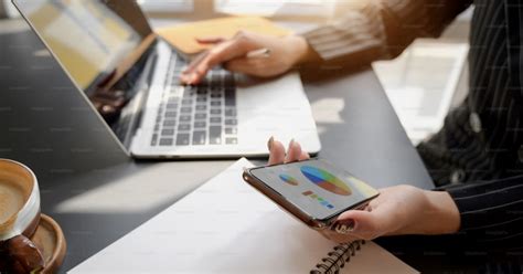 편안한 직장에서 다른 사무용품과 함께 스마트폰으로 비즈니스 차트를 보면서 노트북으로 작업하는 사업가의 측면 모습 사진 Unsplash의 태국 이미지