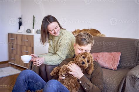 Couple Drinking From Cups Near Sofa In Living Room Red Cat Sleeping On Sofa Cockapoo Running
