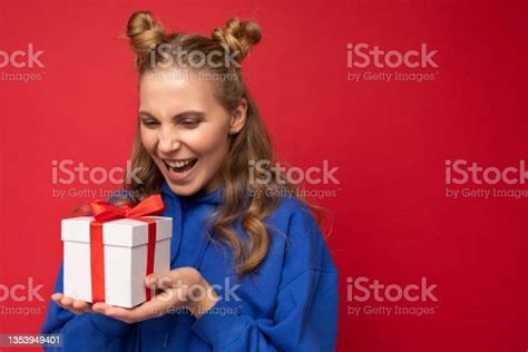 화려한 배경 벽에 고립 된 꽤 긍정적 인 감정적 인 젊은 금발 여성의 사진 사진은 선물 상자를 들고 선물 상자를보고 빨간색 리본으로 선물 상자를보고 화려한 배경 벽을 통해