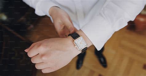 고급 시계를 들고 사업가 세련된 신랑은 흰 셔츠를 입고 시계를 착용하고 아침에 창문 근처에서 부드러운 빛으로 결혼식 전에 준비합니다 시간은 돈이다 사진