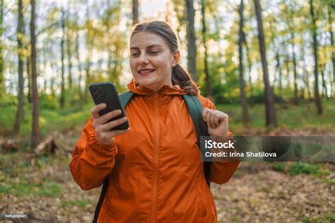 배낭을 메고 따뜻한 옷을 입은 젊은 여성 여행자가 봄 숲에 서서 전화기 화면을 바라보고 있다 등산로에 있는 여자들 숲에 있는 나무 길 폴란드 유럽의 국립 공원 고화질 사진