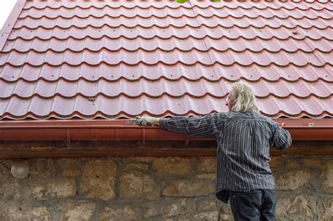 성숙한 남자가 자기 집의 배수로에서 나뭇잎과 부스러기를 치우고 있습니다 빗물 배수구 청소 가득 찬에 대한 스톡 사진 및 기타