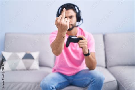 Hispanic Babe Man Playing Video Game Holding Controller Sitting On The Sofa Showing Middle