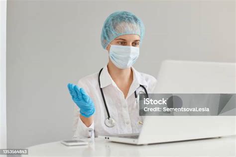 가운 수술용 마스크 의료용 모자 장갑을 착용한 혼란스러운 여성 의사가 노트북 앞에서 포즈를 취하고 무기력한 표정으로 노트북 디스플레이를 보고 병원에서 일하고 있습니다 건강