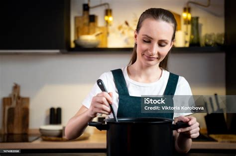 앞치마를 입은 만족스럽고 행복한 젊은 주부 여자는 수프 냄비 숟가락을 들고 냄비에서 수프를 맛본다 가정에있는 사람들의 생활 방식의 개념 서비스에 대한 스톡 사진 및 기타