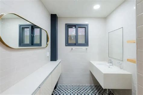 깔끔한 화이트톤의 타일 벽과 패턴 타일 바닥 블랙 프레임의 작은 창과 타원형 거울 화이트 수납장과 세면대가 있는 대학교 센터 화장실 사진