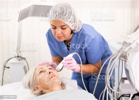 여성 미용사로부터 비수술적 회춘 시술을 받는 노인 여성 스파 센터에서 전기 장치 장비를 사용하여 Rf 얼굴 리프팅 20 24세에