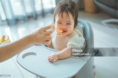 집에서 엄마와 함께 숟가락으로 첫 번째 음식 호박을 먹는 행복한 아시아 아기 소녀 식사 시간 아기의 수유 의자에 앉아서 먹는 어린 소녀 음식을 즐긴다 2명에 대한 스톡 사진