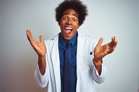 고립된 흰색 배경 위에 코트를 입은 젊은 아프리카 계 미국인 의사가 미친 듯이 축하하고 팔을 들고 눈을 뜨고 흥분한 채 성공을 축하하고 놀랐습니다 우승자 컨셉 건강관리와