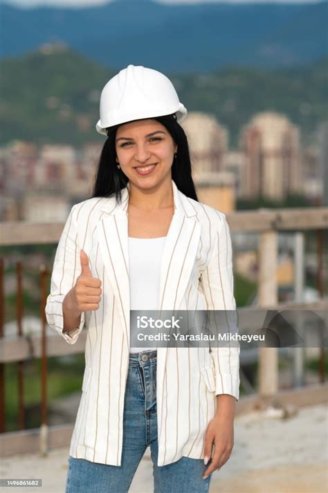 하얀 헬멧을 쓴 젊고 아름다운 웃는 여자가 엄지손가락을 치켜세우는 표지판을 보여준다 건축가 엔지니어 관리자 측량사의 직업 개념 세로 사진 25 29세에 대한 스톡 사진 및