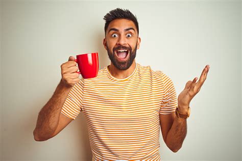 고립된 흰색 배경 위에 서있는 커피의 빨간 컵을 마시는 젊은 인도 남자는 매우 행복하고 흥분 큰 미소와 함께 비명을 지르는 승리를 축하하는 우승자 표정 놀라움에 대한 스톡