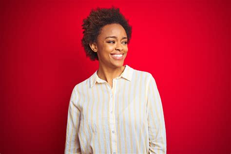 고립된 빨간 배경 위에 아프리카 머리를 가진 젊은 아름다운 아프리카 계 미국인 여성은 얼굴에 미소와 함께 옆으로 찾고 자연스러운 표정 자신감 있게 웃고 있습니다 사람들에