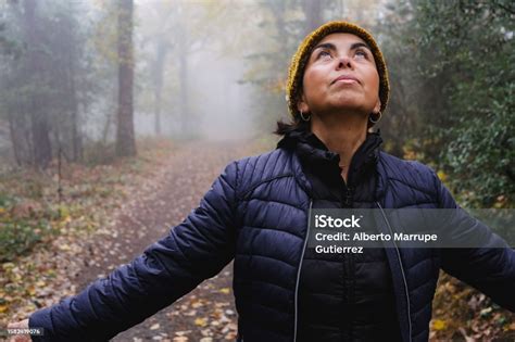 겨울 모자를 쓴 중년 라틴 여성은 땅에 낙엽이 있는 숲 옆 가을 한가운데에 안개가 자욱한 길에서 팔을 벌리고 있다 The Nature Conservancy에 대한 스톡 사진