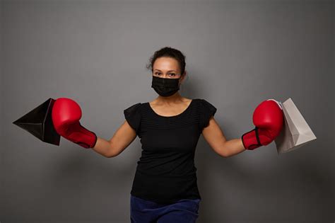 의료 용 마스크 빨간 권투 장갑을 입은 쾌활한 아프리카 여성이 쇼핑 백을 들고 있으며 블랙 프라이데이 광고의 복사 공간이있는 회색 벽 배경에 카메라를 보고 귀엽게 미소를