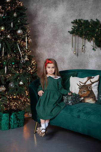 행복한 아이가 선물을 엽니 다 드레스를 입은 어린 소녀는 빛나는 화환 불빛이있는 크리스마스 트리의 소파에 선물로 앉아 있습니다 아름다운 작은 소녀 에 거실 에 자 마스 해피