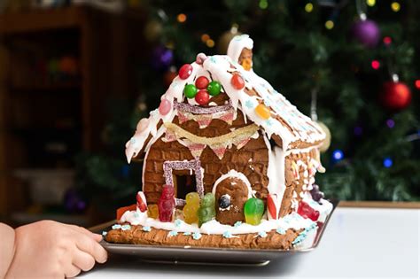 집에서 만든 진저 브레드 하우스는 배경에 흐릿한 크리스마스 트리가있는 착빙 과자 및 젤리 사탕으로 장식되어 있습니다 유아 손이 집을 만진다 12월에 대한 스톡 사진 및 기타