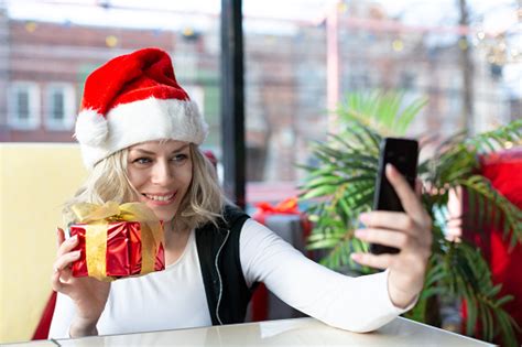 멀리서 크리스마스를 축하합니다 산타 모자를 쓰고 미소 짓는 백인 성인 여성은 카페에서 스마트폰으로 가족과 대화를 나누는 빨간
