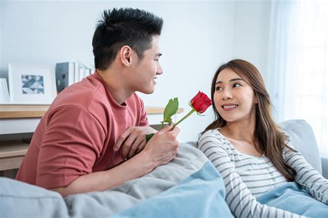 아시아 청년은 놀랍고 아름다운 여자 친구에게 장미 꽃을 줍니다 매력적인 로맨틱 한 새로운 결혼 커플 남성과 여성은 집에서 함께 기념일과 발렌타인 데이를 축하하기 위해 시간을