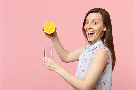 분홍색 파스텔 배경에 고립 된 신선한 잘 익은 오렌지 과일 유리 컵의 절반을 들고 여름 옷에 흥분 충격 젊은 여자 사람들은