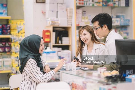 아시아 중국 남성 약사와 함께 약국 카운터에서 히잡을 가진 그녀의 여성 말레이어 고객에게 설명하는 실험실 코트를 가진 아시아 중국 여성 약사 감기와 독감에 대한 스톡 사진