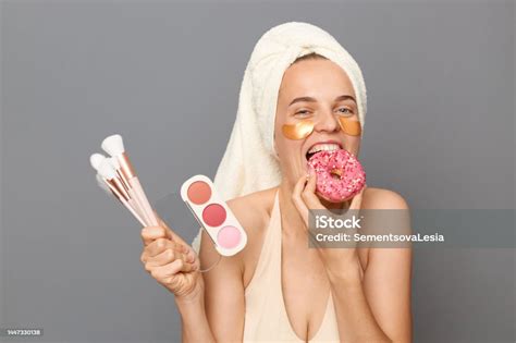 회색 배경 위에 격리된 머리에 상의와 수건을 쓰고 브러시와 아이섀도를 들고 맛있는 도넛을 물고 있는 흥분된 배고픈 웃는 여자의 초상화 30 34세에 대한 스톡 사진 및 기타