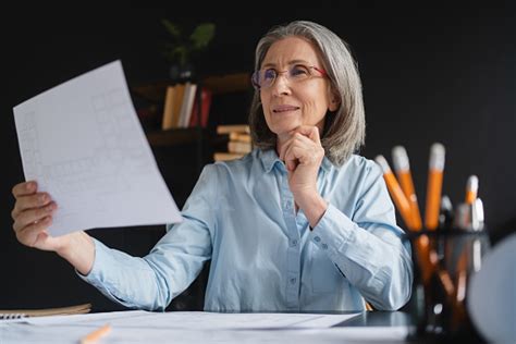 백인 성숙한 여성 건축가가 사무실 책상에 앉아 데스크톱 컴퓨터로 건축 계획을 작업하고 종이 한 장을 들고 창의적인 디자인 프로젝트를 바라보고 있습니다 60 64세에 대한