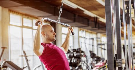 동기 부여되고 집중된 강한 근육질의 스포티 한 피트니스 젊은 대머리 남자가 벤치에 앉아 체육관에서 등 근육을 위해 기계에서 금속 막대를 당기는 측면보기 사진