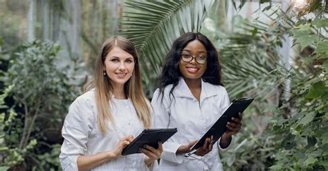 두 명의 젊은 다인종 여성 식물학자 과학자 팀이 열대 식물과 야자수가 있는 온실에서 일하며 클립보드와 태블릿으로 카메라를 향해 웃고 있습니다 사진 Unsplash의