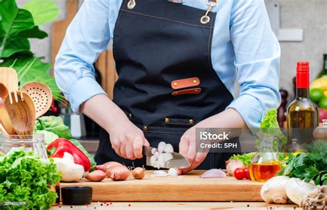 앞치마를 입은 셰프가 나무 테이블 주방용품 야채 허브 요리 재료가 있는 아늑한 주방에서 산토쿠 나이프로 샬롯 양파를 자릅니다 건강한 비건 음식 요리 조리법 음식 블로깅 가정