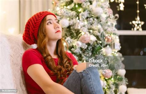 밝은 자연 메이크업을 가진 아름다운 젊은 아시아 여성은 청바지와 함께 빨간 작물 꼭대기에 니트 셔츠를 입고 의자에 앉아 실내에서 새해 장식 배경 크리스마스 트리로 집을 바라