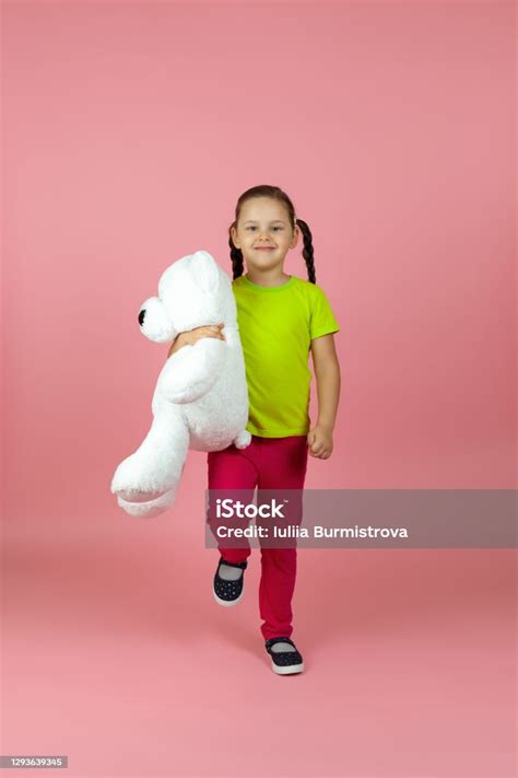 밝은 녹색 티셔츠와 빨간 바지를 입은 전길이의 행복한 피그테일 소녀가 분홍색 배경에 고립된 흰색 테디 베어와 함께 행진합니다 아이에 대한 스톡 사진 및 기타 이미지 Istock
