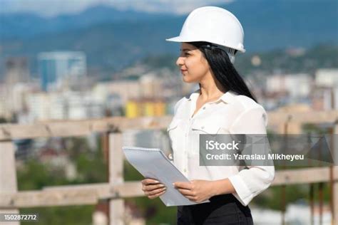 건설 현장에서 안전모를 쓴 젊은 여성 건축가가 종이 태블릿으로 작업하는 동안 시선을 돌린다 건설 현장에서 작업하는 동안 보호 헬멧을 착용한 감독자 공사장 인부에 대한 스톡