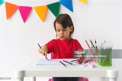 빨간 티셔츠를 입은 어린 아이가 흰 종이에 연필로 그림을 그리고 형형색색의 깃발을 배경으로 미술 용품이 있는 테이블에 앉아 있습니다 어린 시절 창의성 교육의 개념 2 3