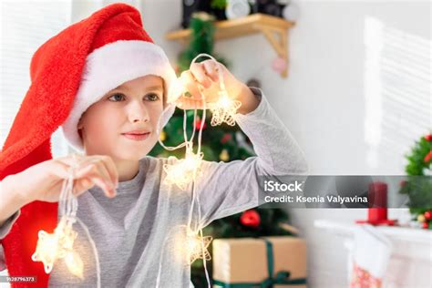 산타 모자를 쓴 어린 소년은 빛나는 화환 불을 손에 들고 크리스마스 축하를 준비합니다이 어린이는 집에서 크리스마스 트리를 장식합니다 기대에 대한 스톡 사진 및 기타 이미지