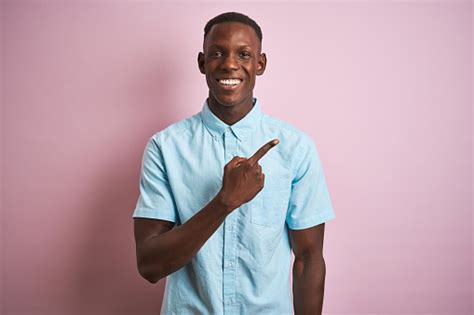 고립된 분홍색 배경 위에 서있는 파란색 캐주얼 셔츠를 입은 아프리카 계 미국인 남성은 얼굴에 행복하고 자연스러운 표정으로 손과 손가락으로 가리키는 얼굴의 미소로 명랑합니다