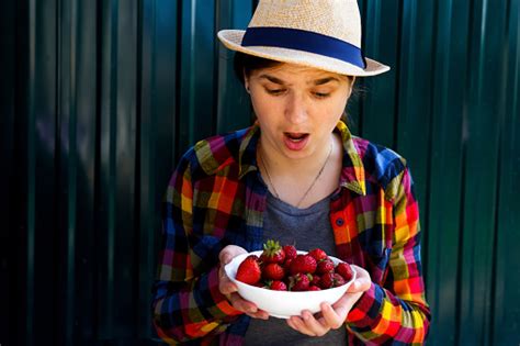 모자와 셔츠를 입은 청녀의 초점을 떼어 내고 미소를 짓고 딸기 한 그릇을 보여 준다 짙은 녹색 강철 배경 여름 음식 과일 여름 배경 제스처처럼 초점이 맞지 않음 25 29