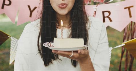 파티 모자를 쓴 세련된 행복한 여자는 생일 케이크 조각에 촛불을 불고 공원에서 분홍색 생일 축하 화환을 배경으로 소원을 빌고 있다 야외 피크닉 파티에서 생일을 축하합니다