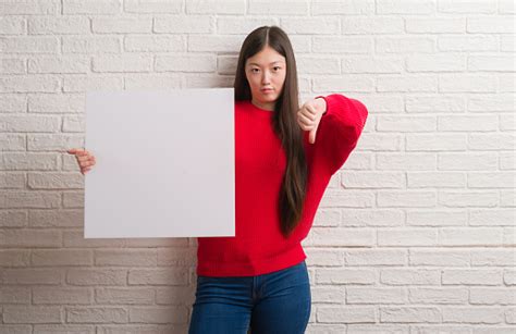 성 난 얼굴 아래로 엄지 싫어 보여주는 음수 부호 거부 개념 배너를 들고 벽돌 벽을 통해 젊은 중국 여자 개념에 대한 스톡 사진 및 기타 이미지 Istock