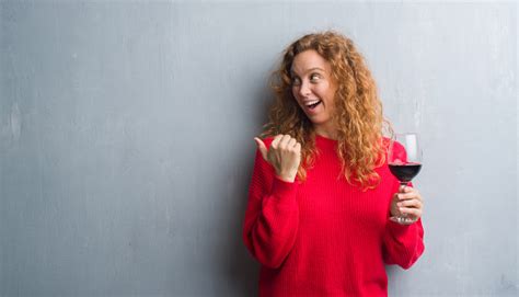 그런 지 회색 벽 고 행복 얼굴 미소와 측면까지 손가락으로 보여주는 와인 한 잔을 마시는 동안 젊은 빨강 머리 여자 가리키기에 대한 스톡 사진 및 기타 이미지 Istock