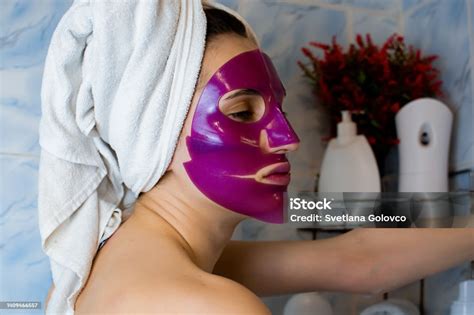 백인 젊은 붉은 머리 여성은 욕실에서 휴식을 취하고 머리를 감싸고있는 목욕 타월을 착용하고 얼굴에 분홍색 점토 얼굴 마스크가있는 거품 욕조에 누워 손톱 매니큐어를 즐깁니다