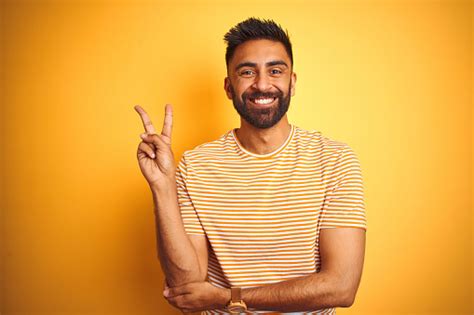고립된 노란색 배경 위에 서있는 티셔츠를 입은 젊은 인도 남자가 손가락으로 승리 표지판을하고있는 카메라에 행복한 얼굴 윙크와