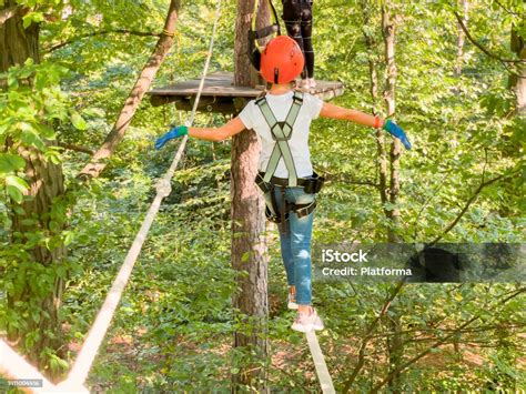 해피 등반 어드벤처 파크에서 활동을 즐기는 십대 소녀는 여름날 안전 장비와 헬멧 사이의 밧줄 다리를 따라 걷습니다 나무 숲에있는 로프 공원 모험 공원이 닫힙니다 모험심
