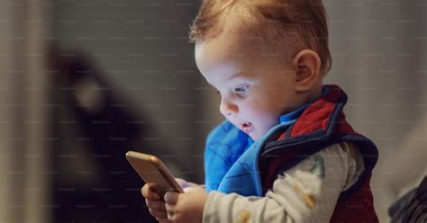 사랑스러운 놀란 작은 금발 소년은 어머니의 무릎에 앉아 스마트 폰을 들고 있습니다 그는 인터넷에서 서핑을하고 있습니다 사진 Unsplash의 세르비아 이미지