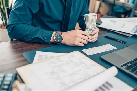 비즈니스 맨은 사무실에서 책상에 흰색 빈 은행 수표 책과 달러 지폐 동전 노트북 및 그래프 차트에 서명 손으로 수표 급여 급여 개념으로 지불합니다 Po에 대한 스톡 사진 및