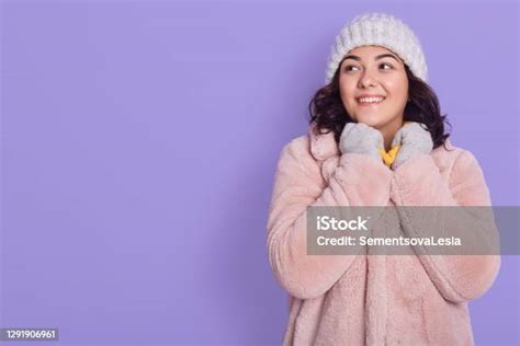 따뜻한 모피 코트와 모자를 쓰고 파스텔 라일락 색상 배경에 고립 된 옆으로 찾고 긍정적 인 감정을 가지고 프로모션공간을 복사