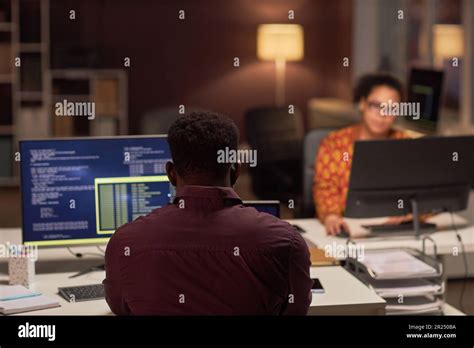 Rear View Of Programmer Sitting At His Workplace In Front Of Computer And Typing Codes In Office