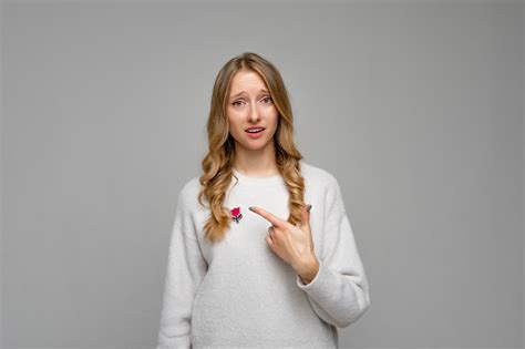 충격을 받고 걱정스러운 젊은 금발 의 여자는 자신을 손가락으로 가리키고 찡그린 얼굴을 찌푸리고 화가 났으며 흰색 스웨터를 입고 회색 배경 위에 서 있습니다 25 29세에
