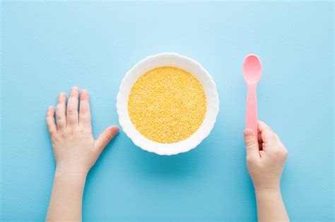 분홍색 플라스틱 숟가락을 들고있는 아기 손 연한 파란색 테이블 배경에 마른 노란색 콘밀 죽이있는 흰색 그릇 파스텔 색상 근접 촬영 시점 촬영 어린이 건강 식품 하향식 보기