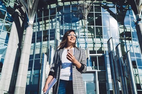 스마트폰으로 도시 환경을 걷는 세련된 옷을 입은 쾌활한 힙스터 소녀 도시의 화창한 날과 현대 건축물에서 자유 시간을 즐기는 휴대폰으로 알림을 확인하는 행복한 젊은 여성 20