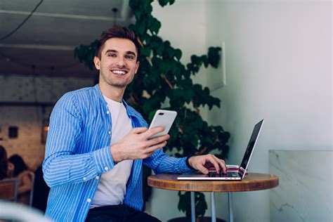 원격 작업을 위해 현대적인 노트북 컴퓨터를 들고 테이블에 앉아 카메라를 향해 웃고 있는 쾌활한 남성 프로그래머의 절반 길이 초상화 넷북과 핸드폰 장치를 사용하는 행복한 백인